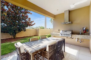 Cozinha externa com churrasqueira, mesa de jantar e cadeiras, cercada por um jardim e uma parede de tijolos.