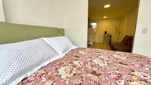 Uma cama com cabeceira acolchoada e edredom floral, uma cadeira marrom e uma mesinha com uma xícara e uma garrafa.