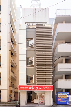 Um edifício de vários andares com design moderno, destacando-se por uma fachada vertical com ripas e uma entrada no nível da rua com uma placa vermelha.