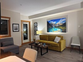Uma sala de estar com sofá, mesa de centro, poltrona, luminária, quadros emoldurados e uma grande pintura de paisagem.