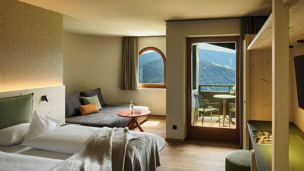 Quarto de hotel com duas camas, uma mesinha com garrafa e copos, um sofá com almofadas, uma janela com vista para montanhas e uma varanda com mesa e cadeiras.