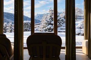 Vista da janela com uma cadeira em primeiro plano, com paisagem nevada de árvores e montanhas.