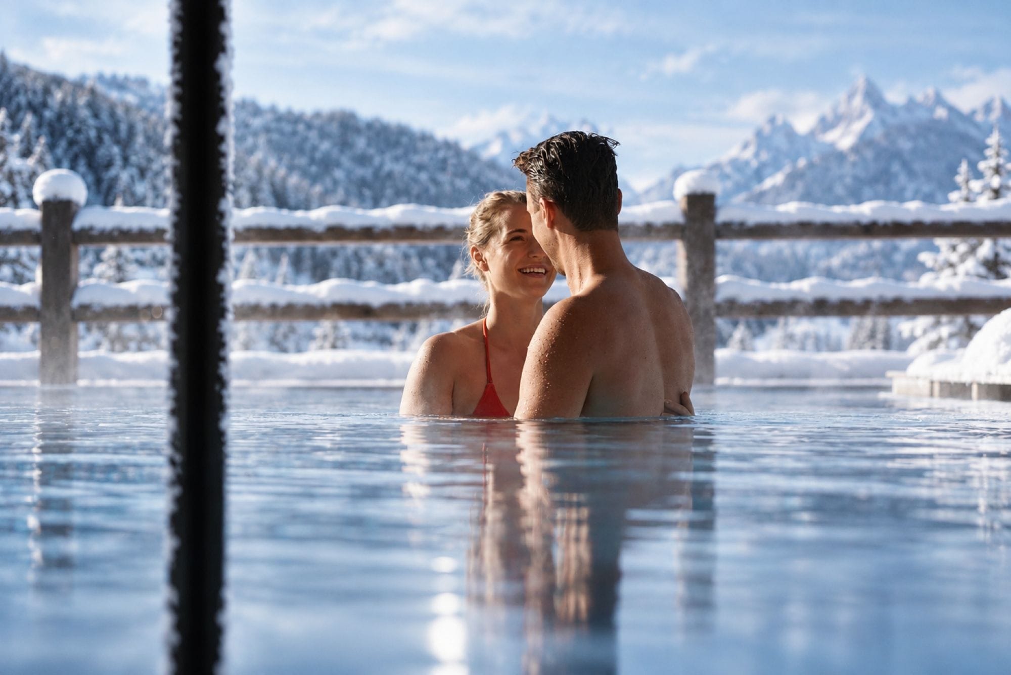Um casal em uma banheira de hidromassagem com montanhas nevadas ao fundo.
