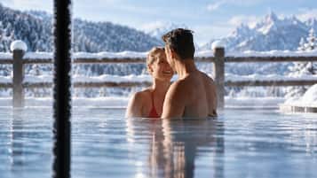 Um casal em uma banheira de hidromassagem com montanhas nevadas ao fundo.