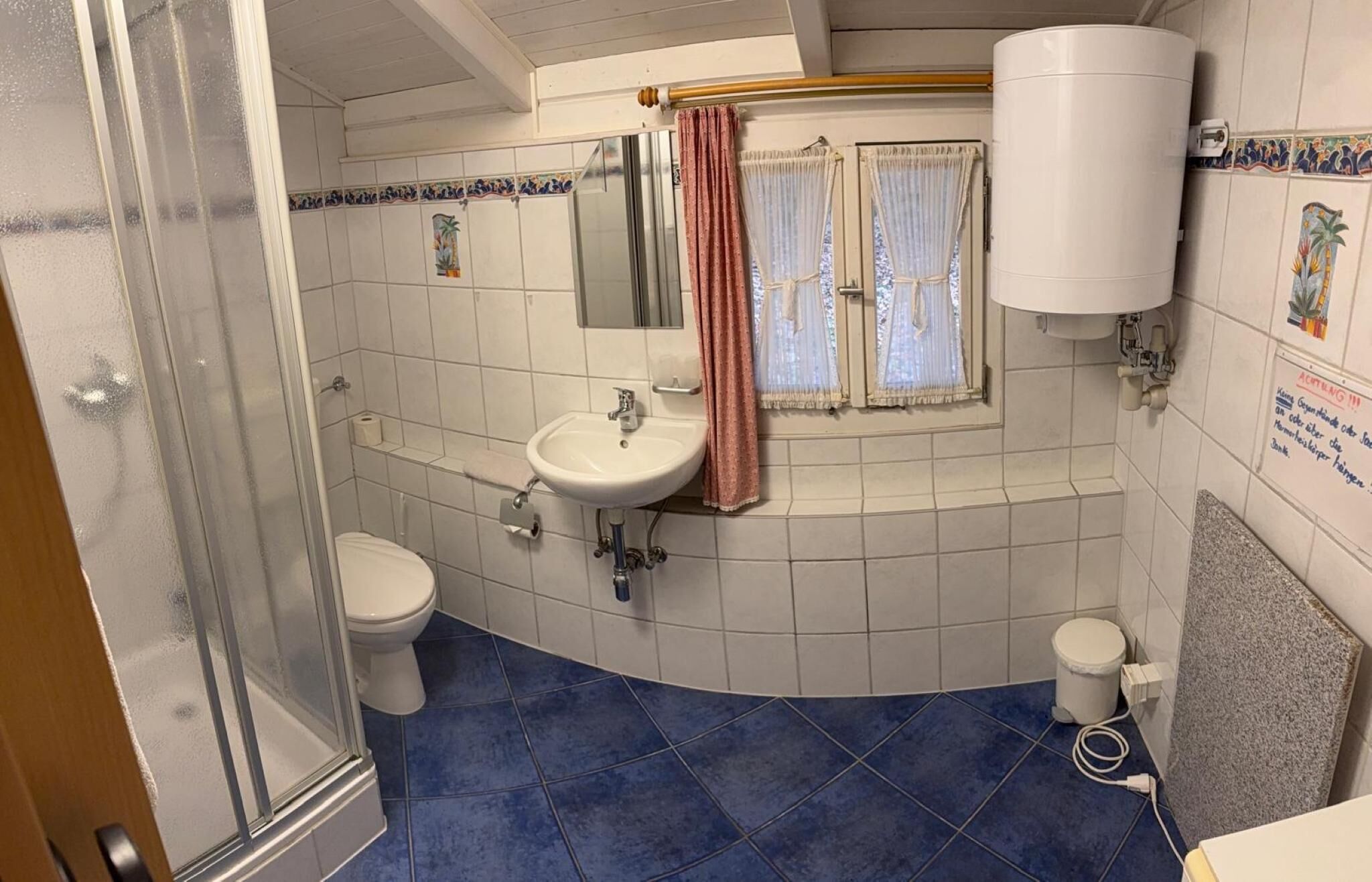 Casa de banho com chuveiro, sanita, lavatório e janela com cortinas.