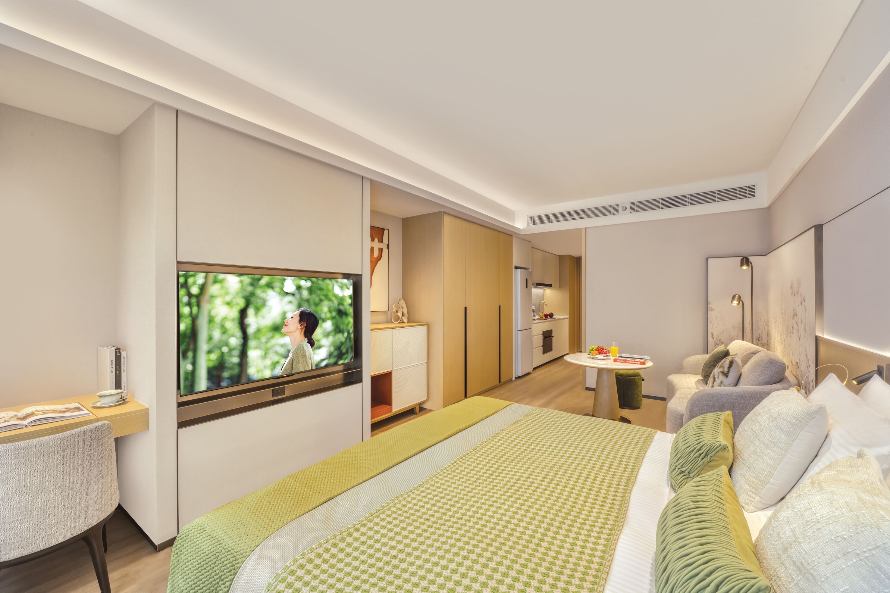 Quarto de hotel moderno com cama, televisão, escrivaninha e uma pequena cozinha equipada.