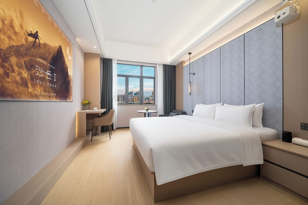 Quarto de hotel com uma cama grande, uma escrivaninha, uma cadeira, uma mesinha e uma janela com vista para a cidade.