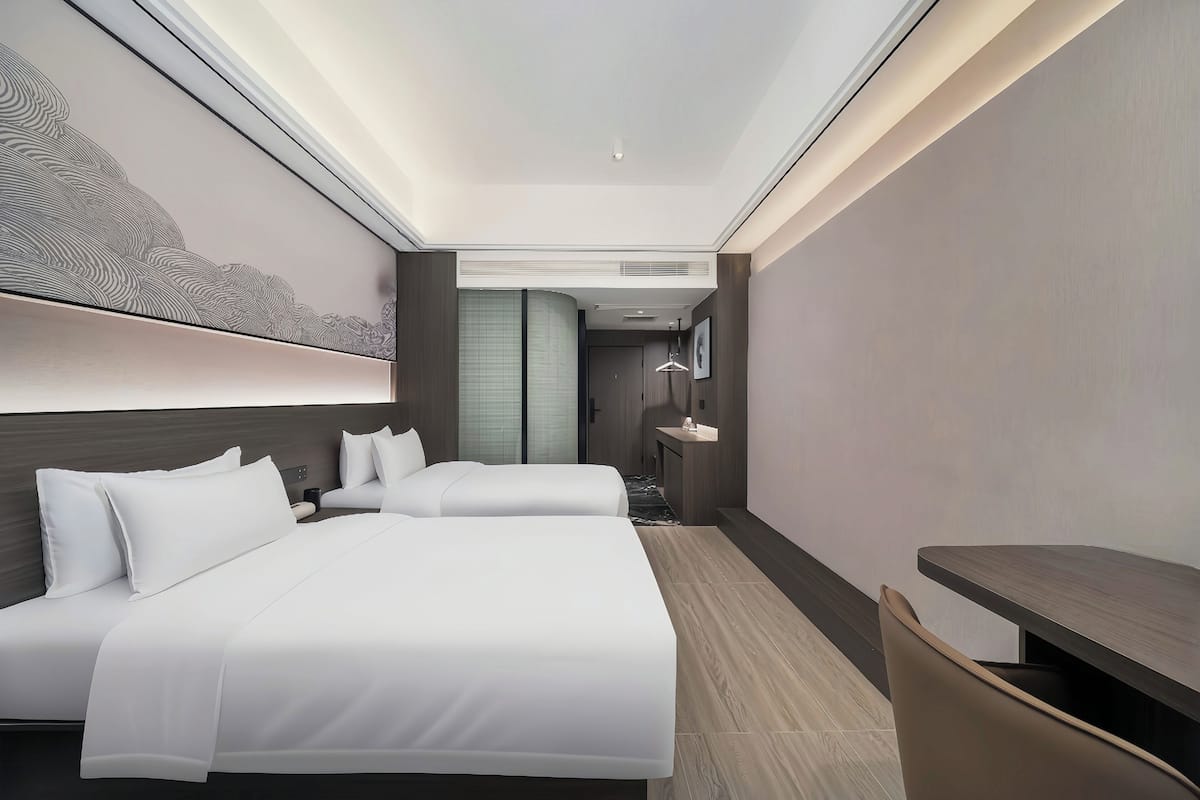 Quarto de hotel com uma cama grande, uma escrivaninha e um banheiro visível através de uma porta de vidro.