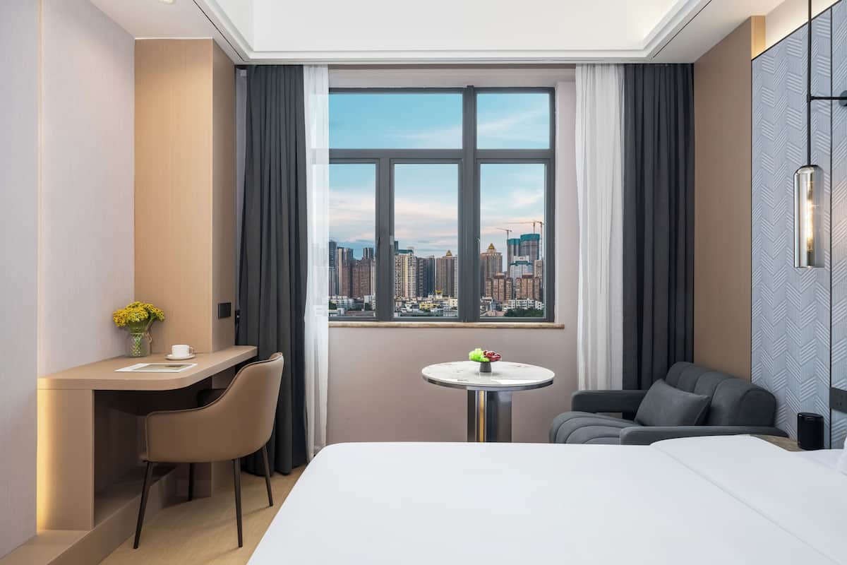 Quarto de hotel com cama, escrivaninha, cadeira, mesa redonda pequena e vista para a paisagem urbana através de grandes janelas.