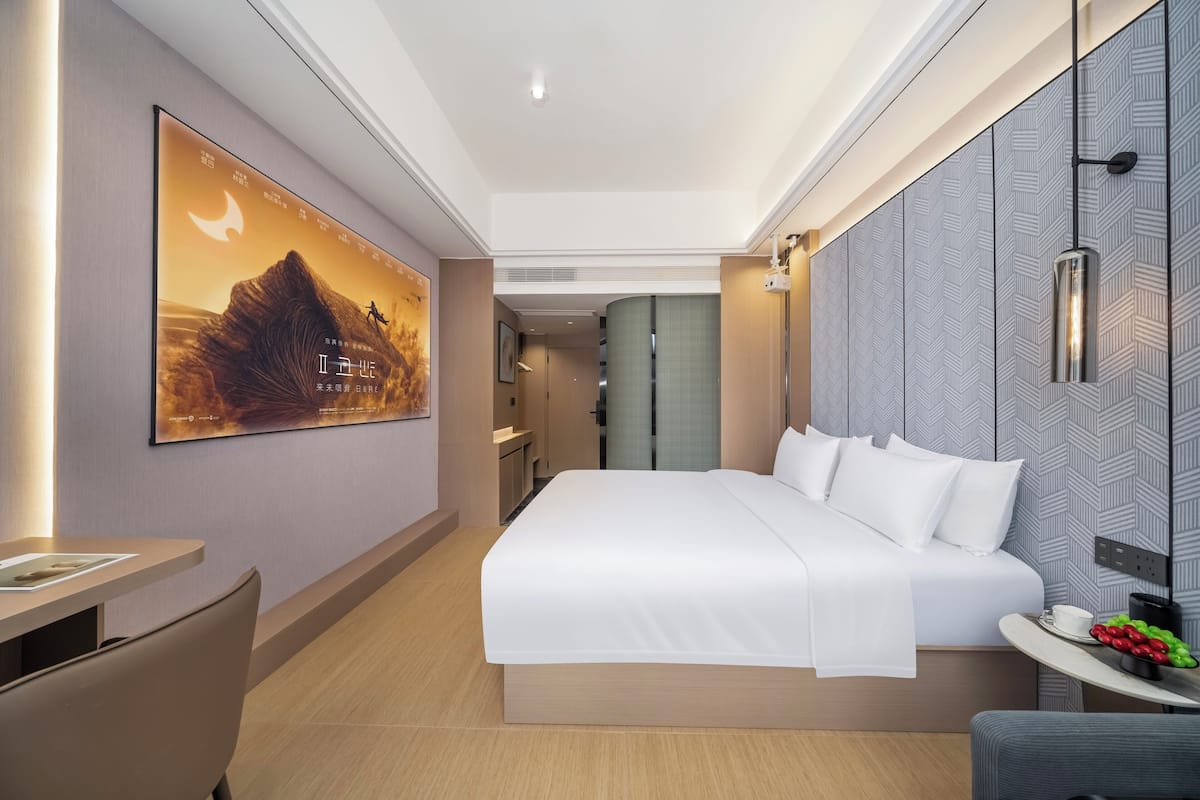 Quarto de hotel com uma cama grande, uma escrivaninha, uma cadeira, uma mesinha com uma chávena e fruta, e um quadro na parede.