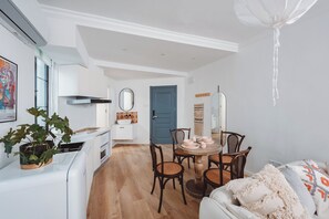 Uma cozinha moderna com armários brancos, um espaço de refeições com uma mesa redonda e cadeiras, e uma porta azul ao fundo.