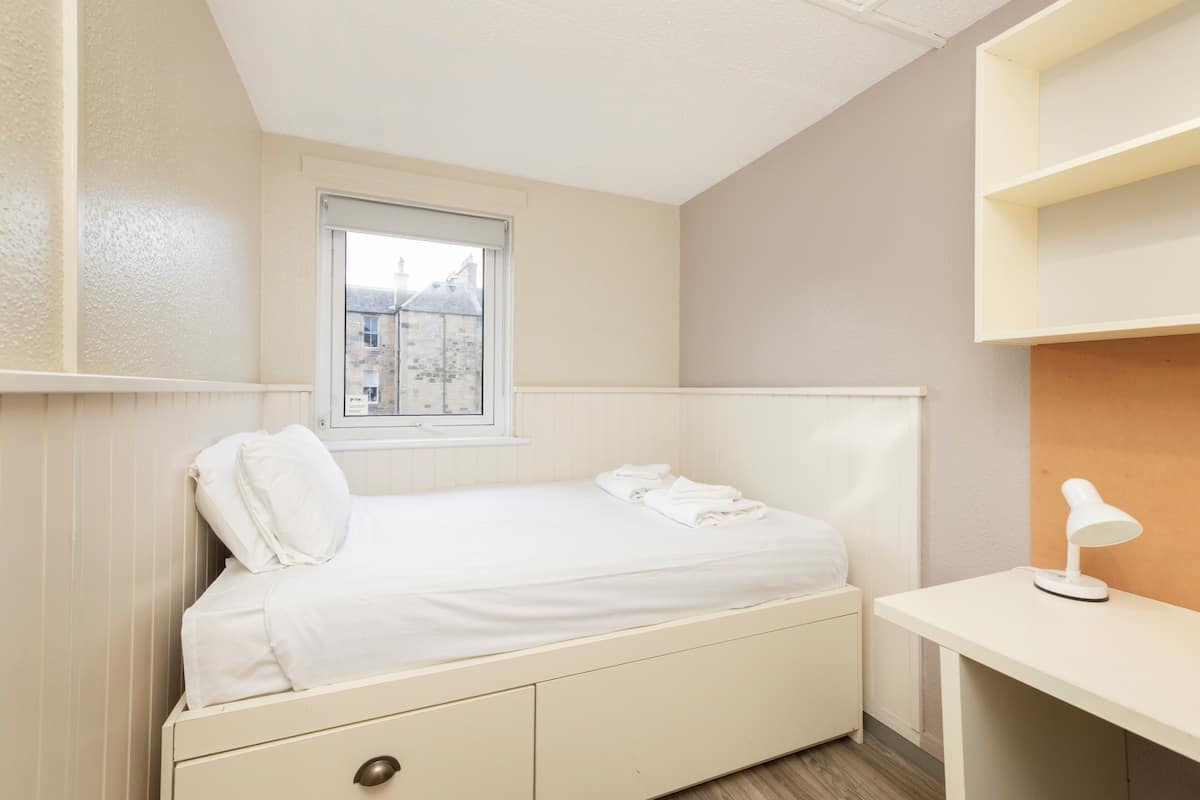 Quarto com cama, escrivaninha, janela e vista para prédios.