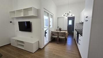 Cozinha moderna com armários brancos, área de jantar com mesa e cadeiras, e TV fixada na parede.