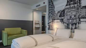 Quarto de hotel com uma cama, uma poltrona verde, um mural na parede retratando uma paisagem urbana e uma porta que dá para outro cômodo.