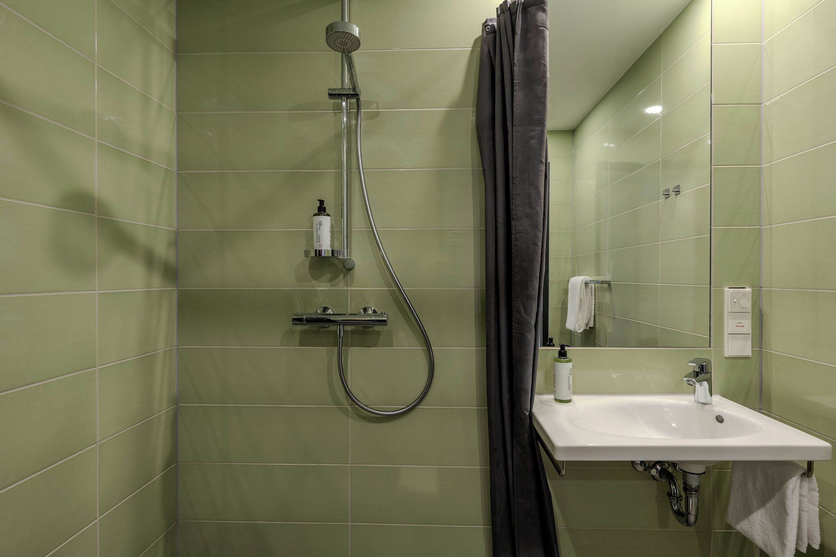 Casa de banho com azulejos verdes, chuveiro, pia e um porta-toalhas.