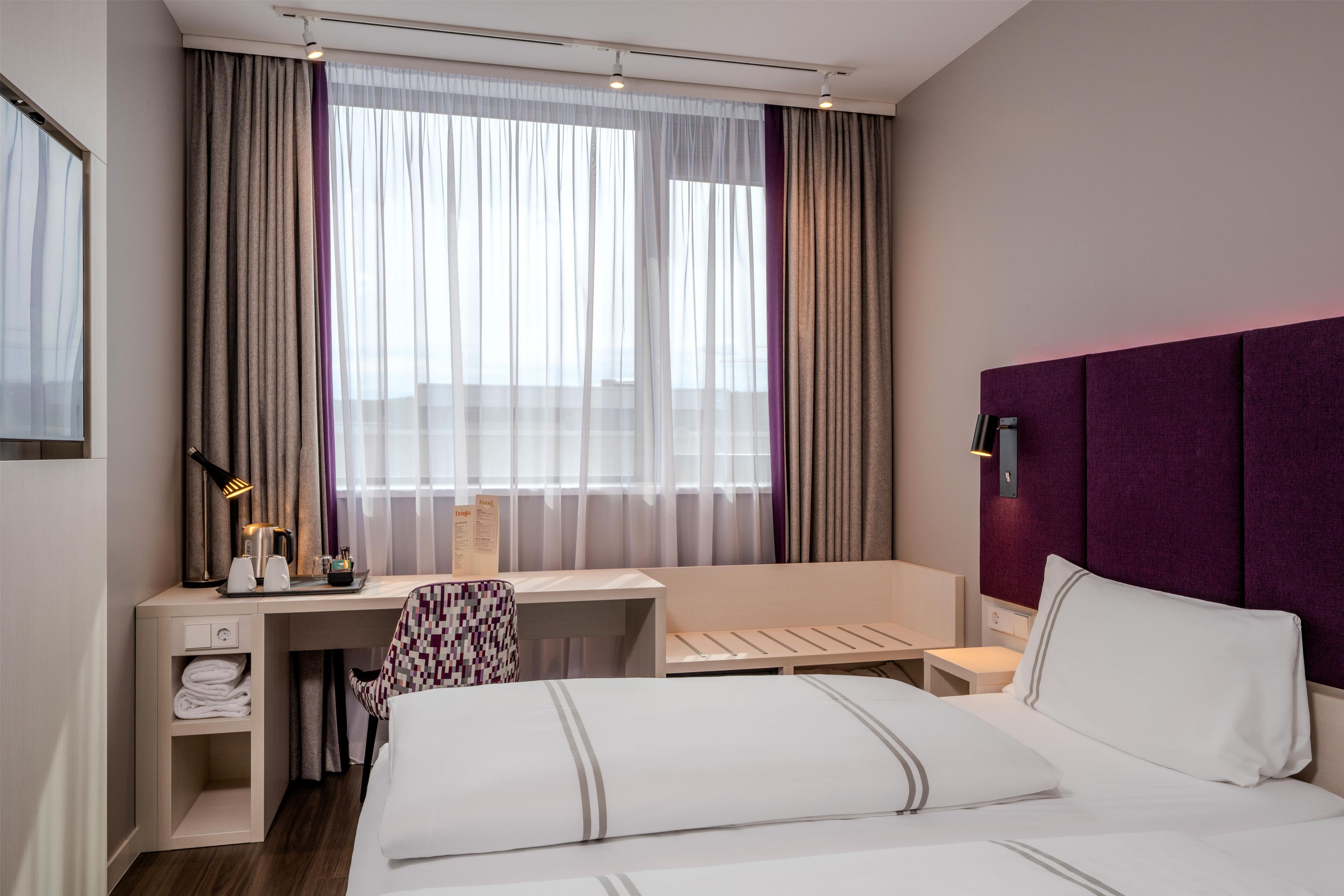 Quarto de hotel com cama, escrivaninha, cadeira, mesa de cabeceira e janela com cortinas.
