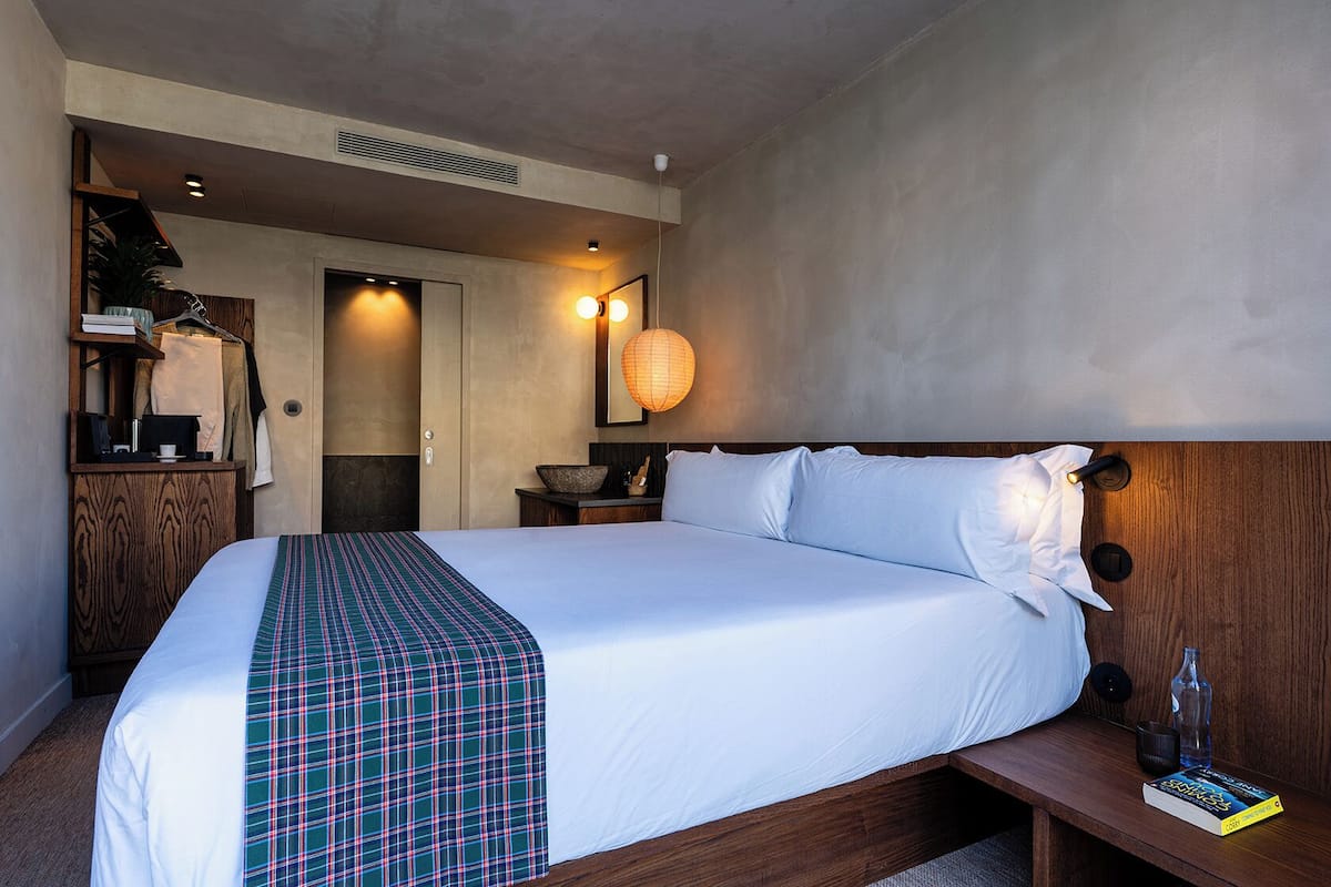 Quarto de hotel com cama grande, mesas de cabeceira, espelho, luminária na parede e porta para outro cômodo.