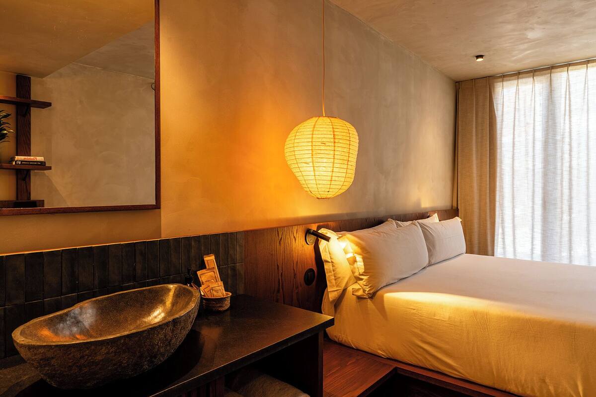 Quarto de hotel moderno com uma cama grande, uma bacia de pedra, uma luminária de parede e um espelho.