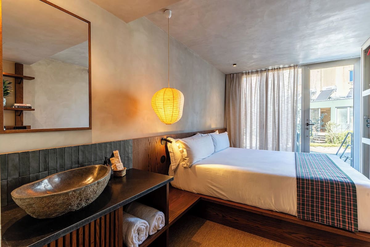 Quarto de hotel com uma cama grande, uma mesa de cabeceira de madeira com lavatório embutido, um espelho e uma janela com cortinas.