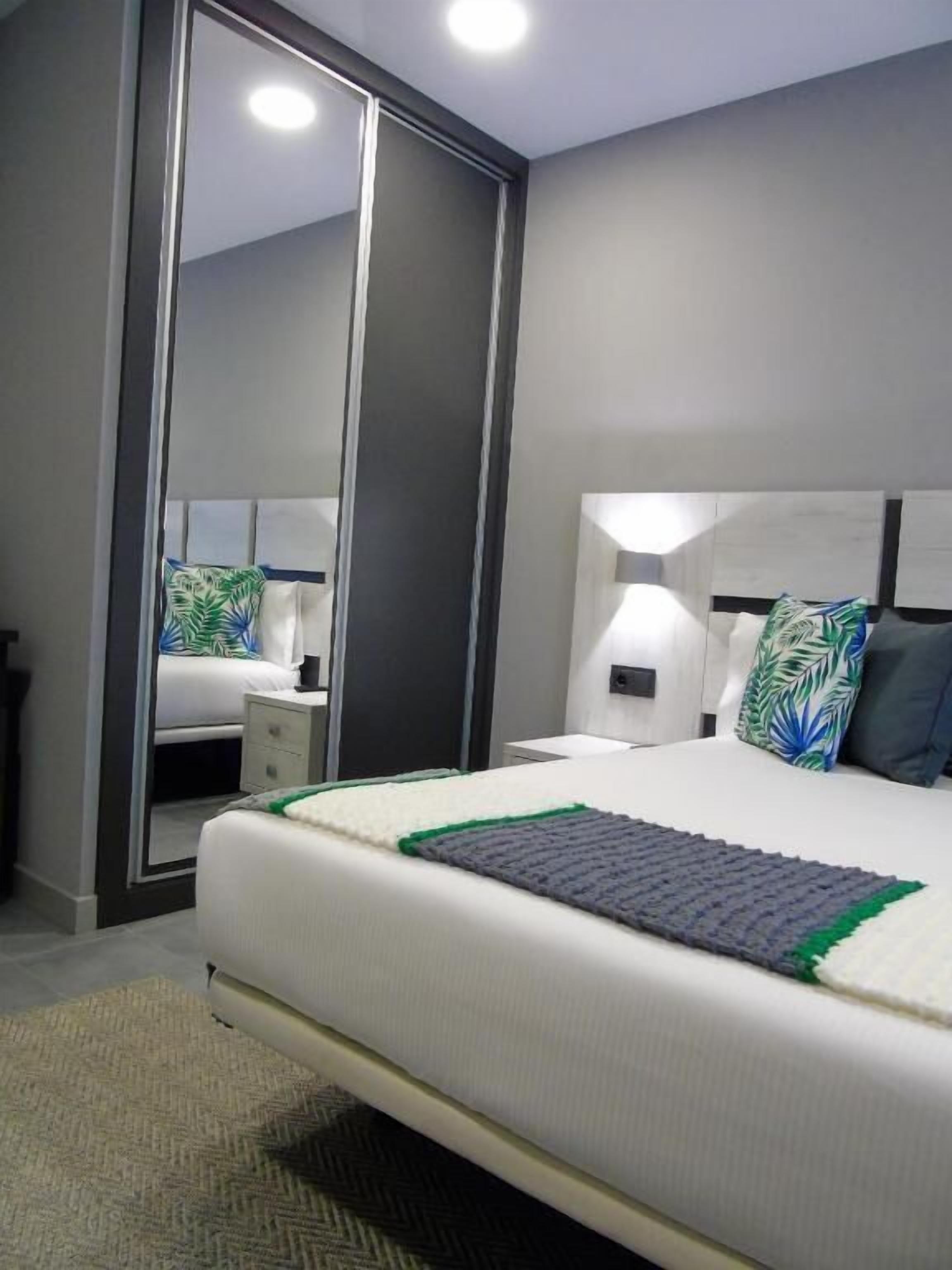 Quarto de hotel moderno com uma cama grande, mesa de cabeceira, espelho e luminária fixada na parede.
