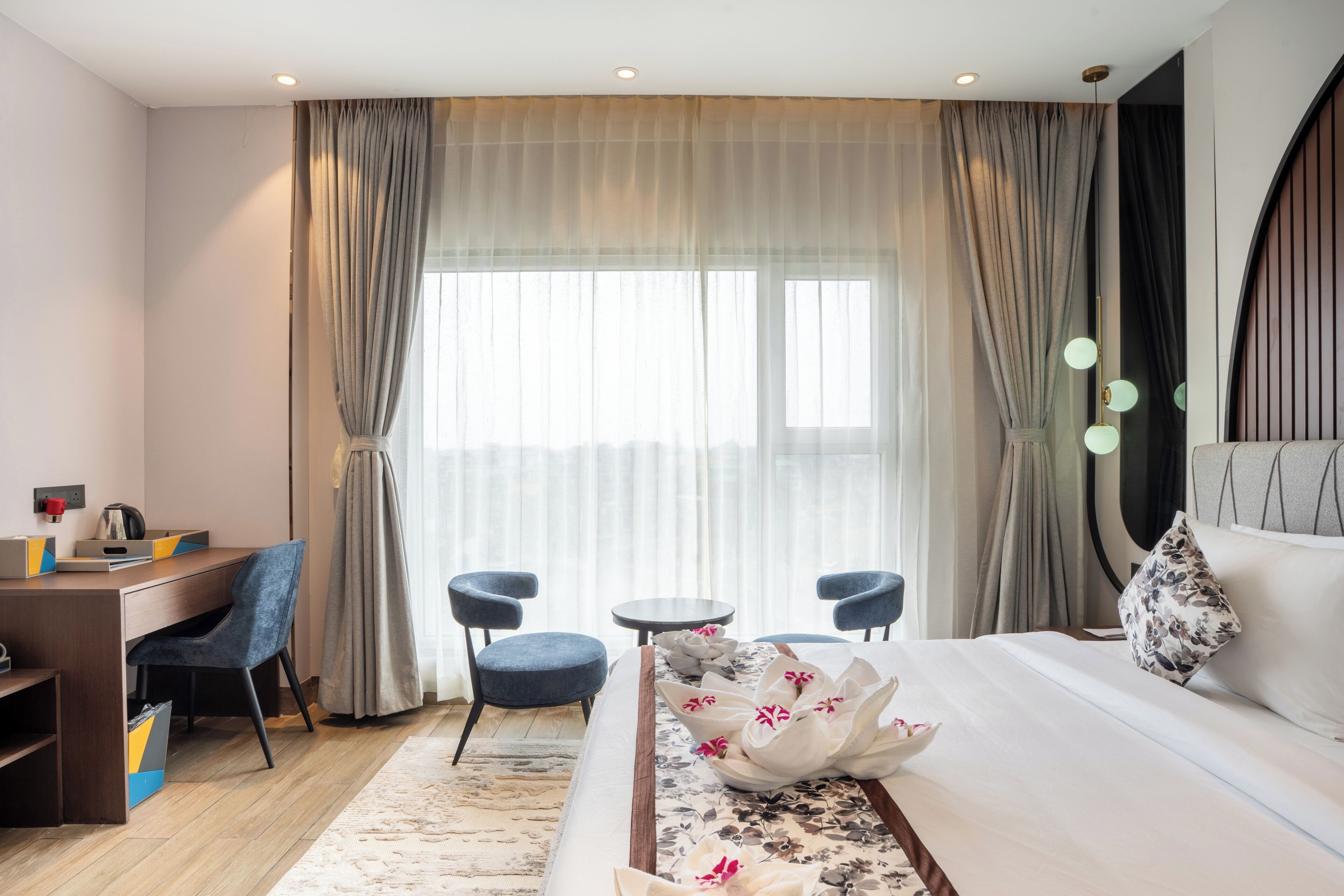 Quarto de hotel com cama, escrivaninha, cadeiras e vista da cidade por grandes janelas.