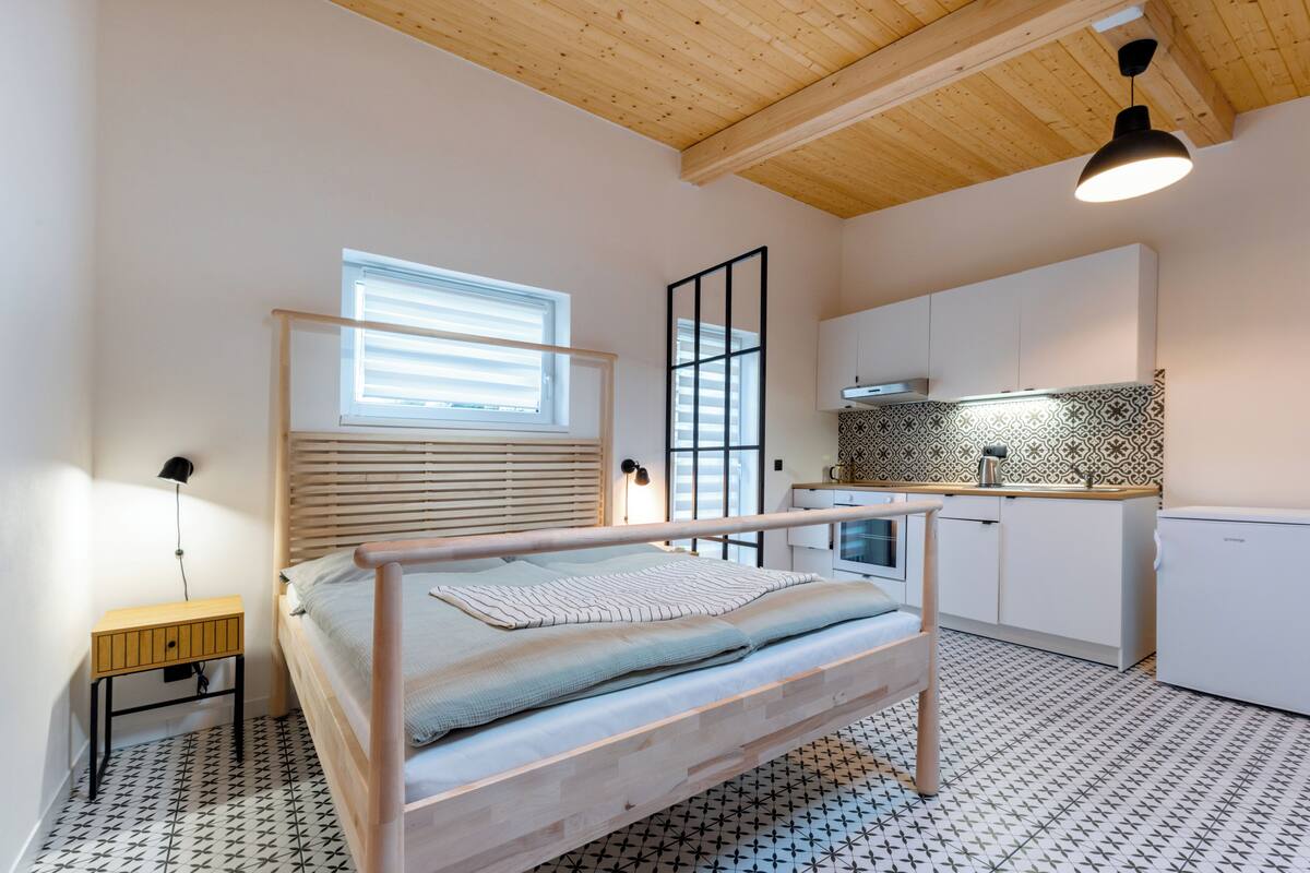 Quarto com cama de madeira, mesa de cabeceira pequena, janela com persianas e área de cozinha com armários brancos e painel decorativo.