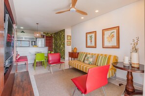 Uma sala de estar com sofá, cadeiras, mesa de jantar, ventilador de teto e cozinha ao fundo.