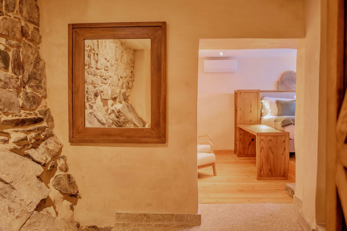Paredes de pedra com um quadro emoldurado, um banco de madeira e uma cama com almofadas em um quarto.