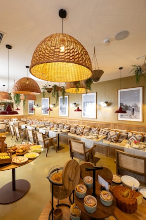 Uma área de refeições com luminárias pendentes de vime, assentos acolchoados e uma mesa de buffet com diversos pratos.