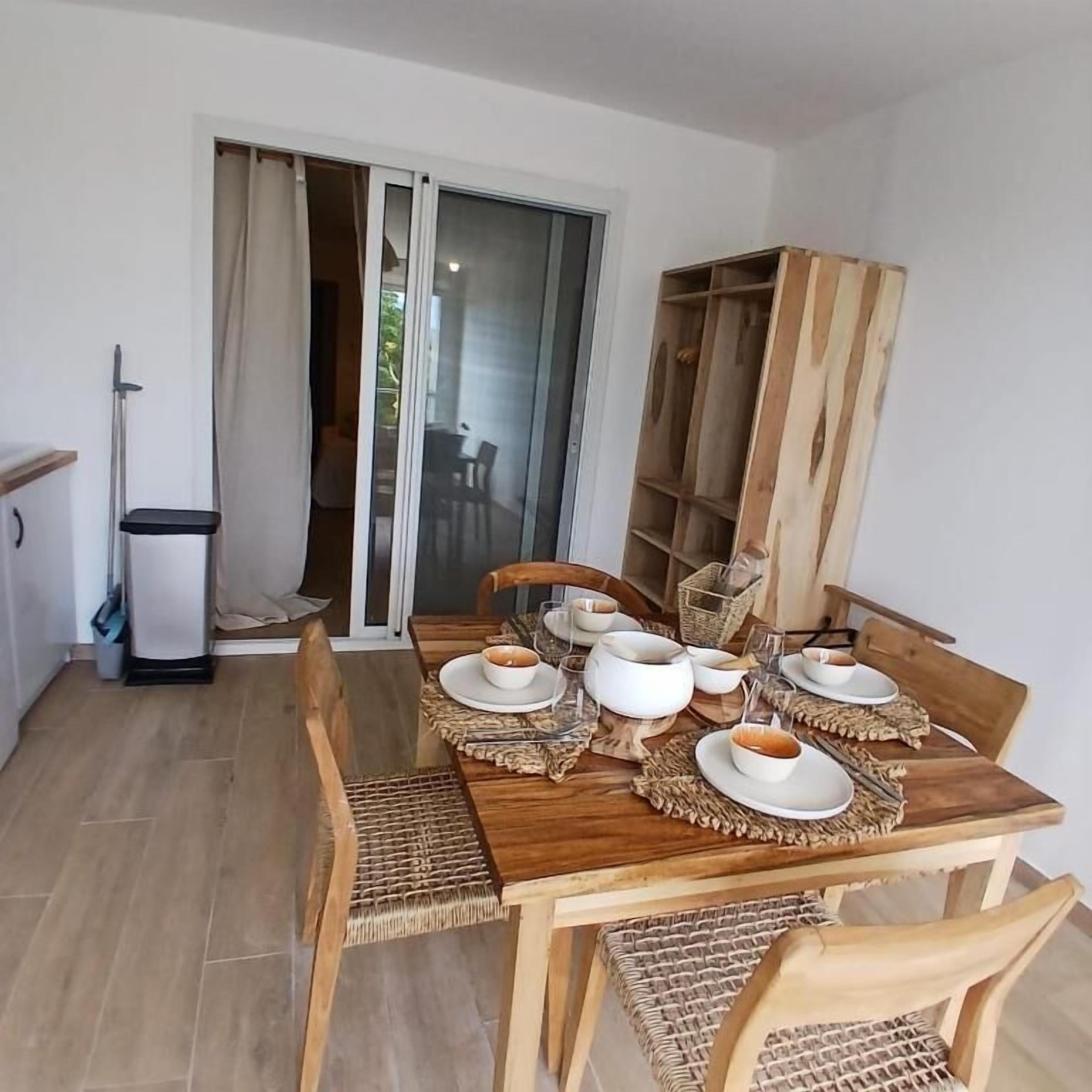 Uma área de refeições com mesa de madeira posta para quatro pessoas, uma porta de vidro deslizante que dá para uma área externa e uma estante de madeira.