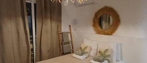 Quarto com cama, mesa de cabeceira com bandeja, luminária pendente, espelho e janela com cortinas.