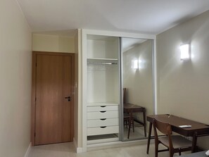 Quarto com porta de madeira, roupeiro branco com gavetas, porta espelhada do roupeiro, pequena mesa com cadeira e escrivaninha com cadeira.