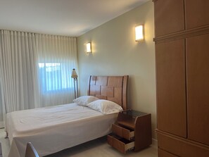 Quarto com cama, mesa de cabeceira com gavetas, luminária e guarda-roupa.