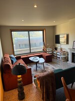 Uma sala de estar moderna com sofá, mesinha, luminária, lareira e uma grande janela com vista para um edifício.