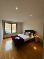 Quarto com uma janela grande, uma cama com almofadas, um abajur e piso de madeira.