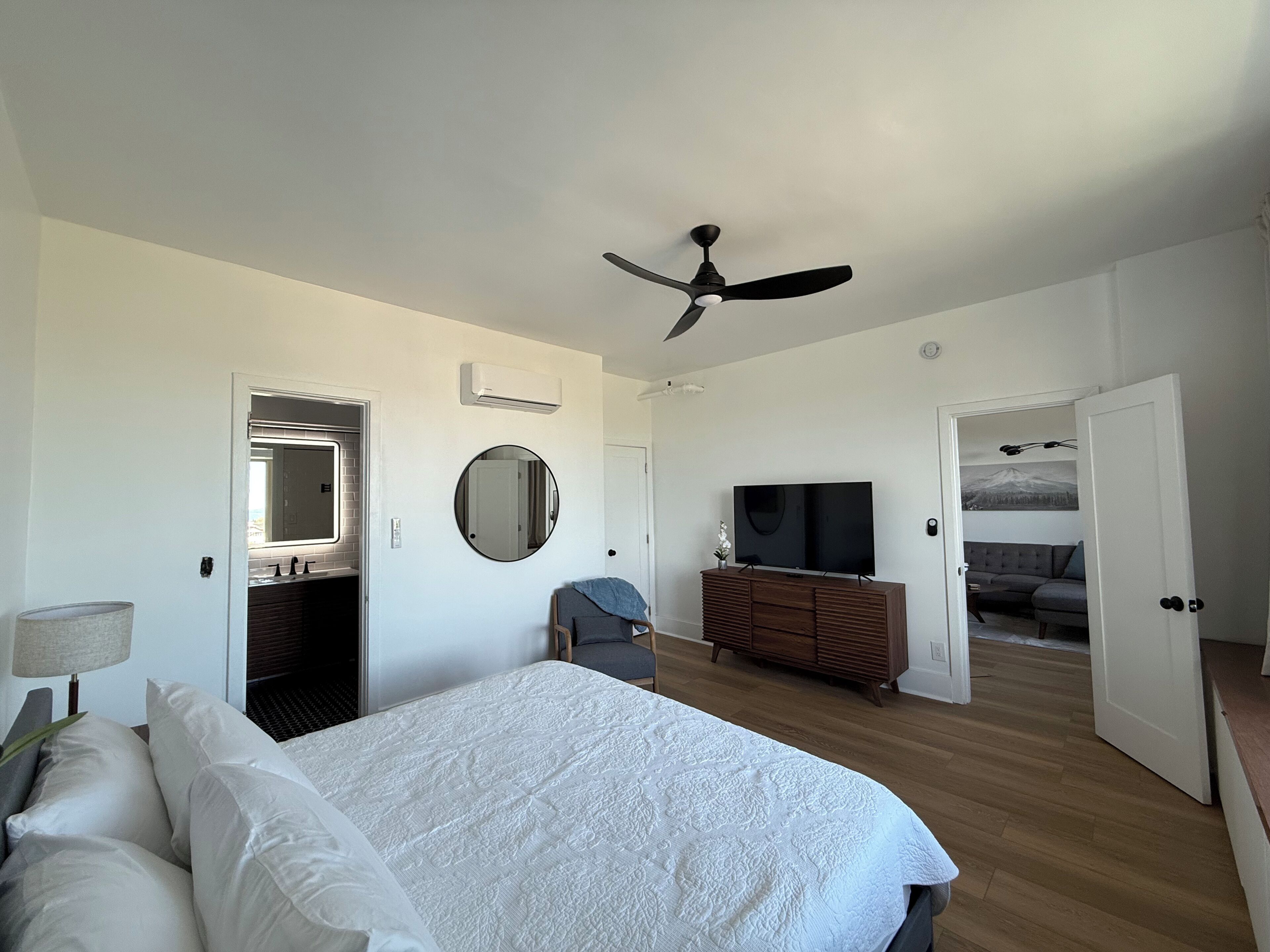 Quarto com cama, ventoinha no teto, cômoda, televisão e porta que dá para uma sala de estar.