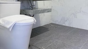 Casa de banho com parede de mármore, lavatório, espelho, secador de cabelo e um contentor de lixo branco.
