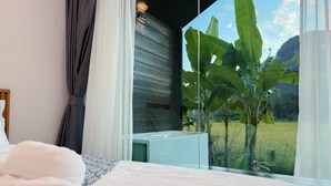 Quarto com uma janela grande, uma cama, uma mesinha de cabeceira e vista para uma paisagem verde.