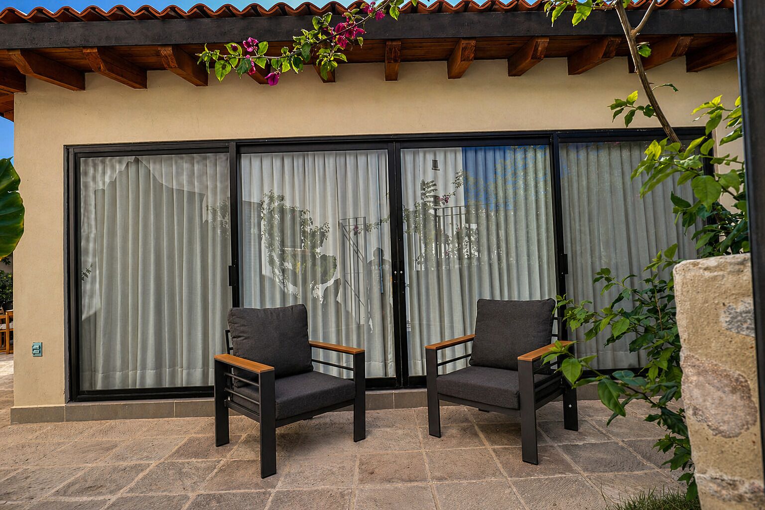 Um pátio com duas cadeiras, uma pérgola e uma porta de correr com cortina decorativa.
