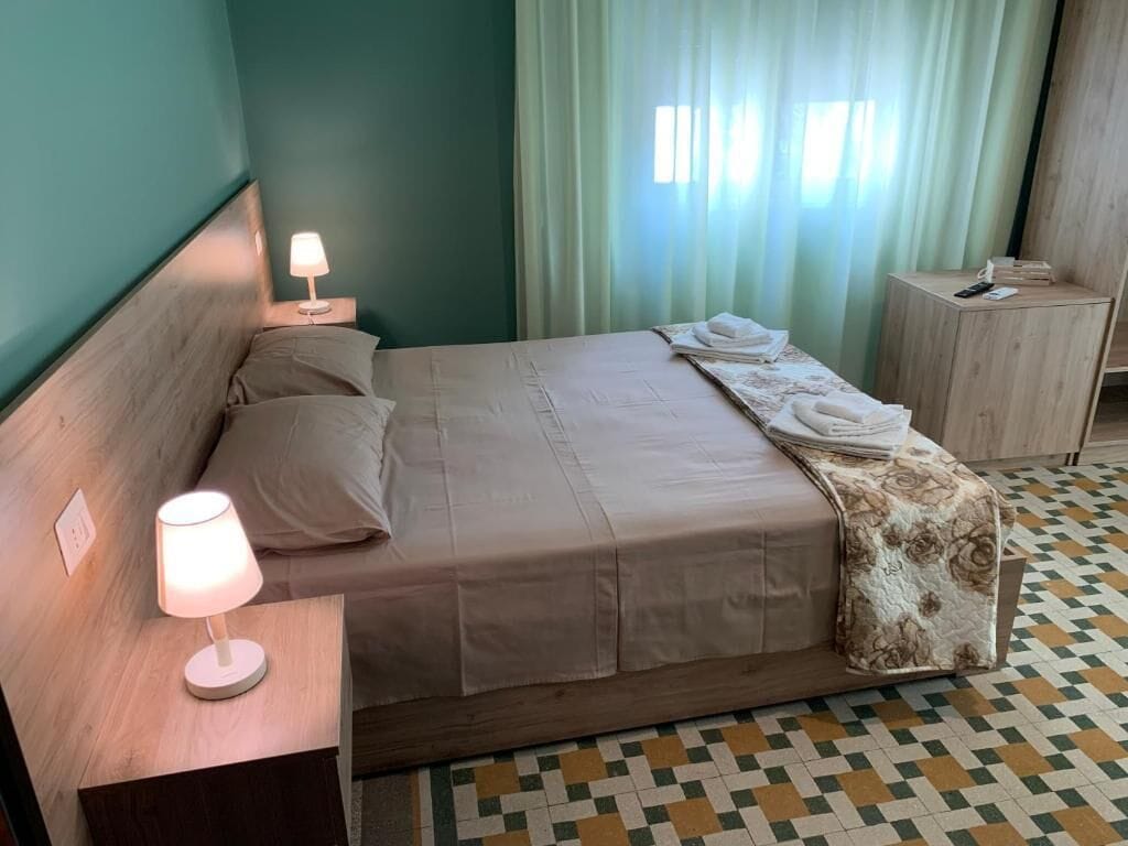 Cama bem arrumada com edredom branco e almofadas, luminária de cabeceira, mesa de cabeceira de madeira e manta decorativa.