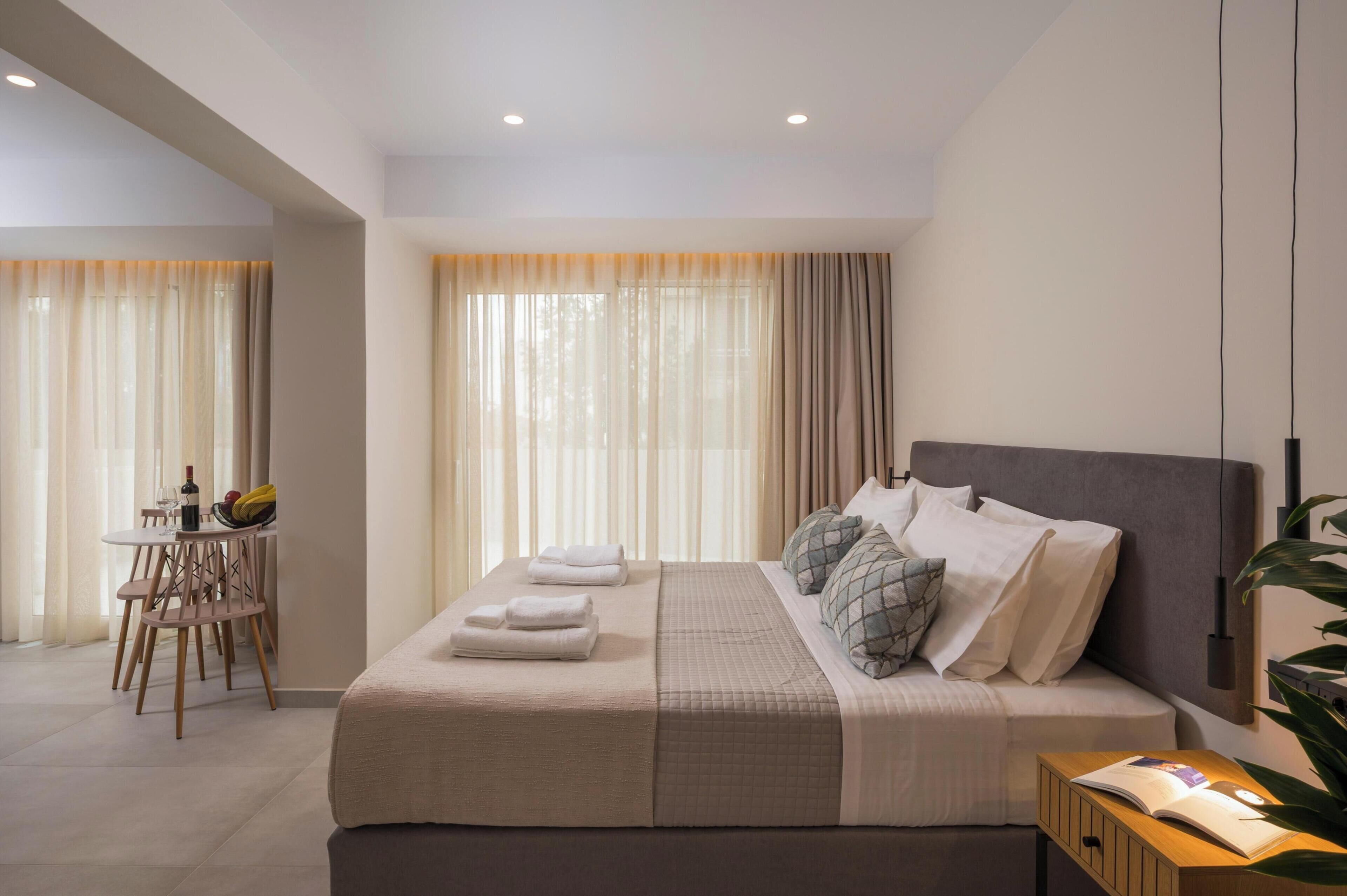 Quarto de hotel com cama, mesa de cabeceira, cortinas e área de refeições com cadeiras e uma mesa.