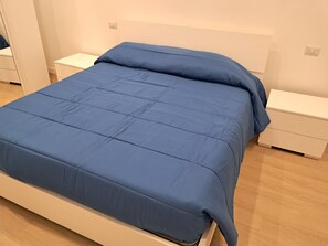 Uma cama com colchão e almofadas azuis, uma mesa de cabeceira e um criado-mudo.