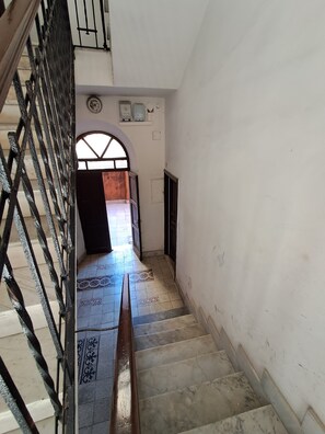Uma escadaria com corrimão de metal, que leva a um quarto com soalho em azulejos e uma porta.