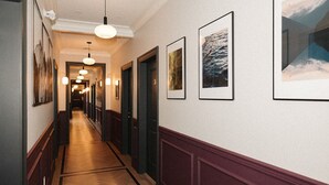 Corredor de hotel com obras de arte emolduradas nas paredes, piso de madeira e iluminação pendente.