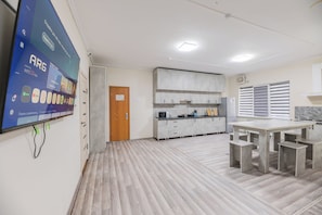 Um quarto espaçoso com uma grande televisão de tela plana na parede, uma porta de madeira, uma cozinha com bancada de mármore e uma mesa de jantar com bancos.