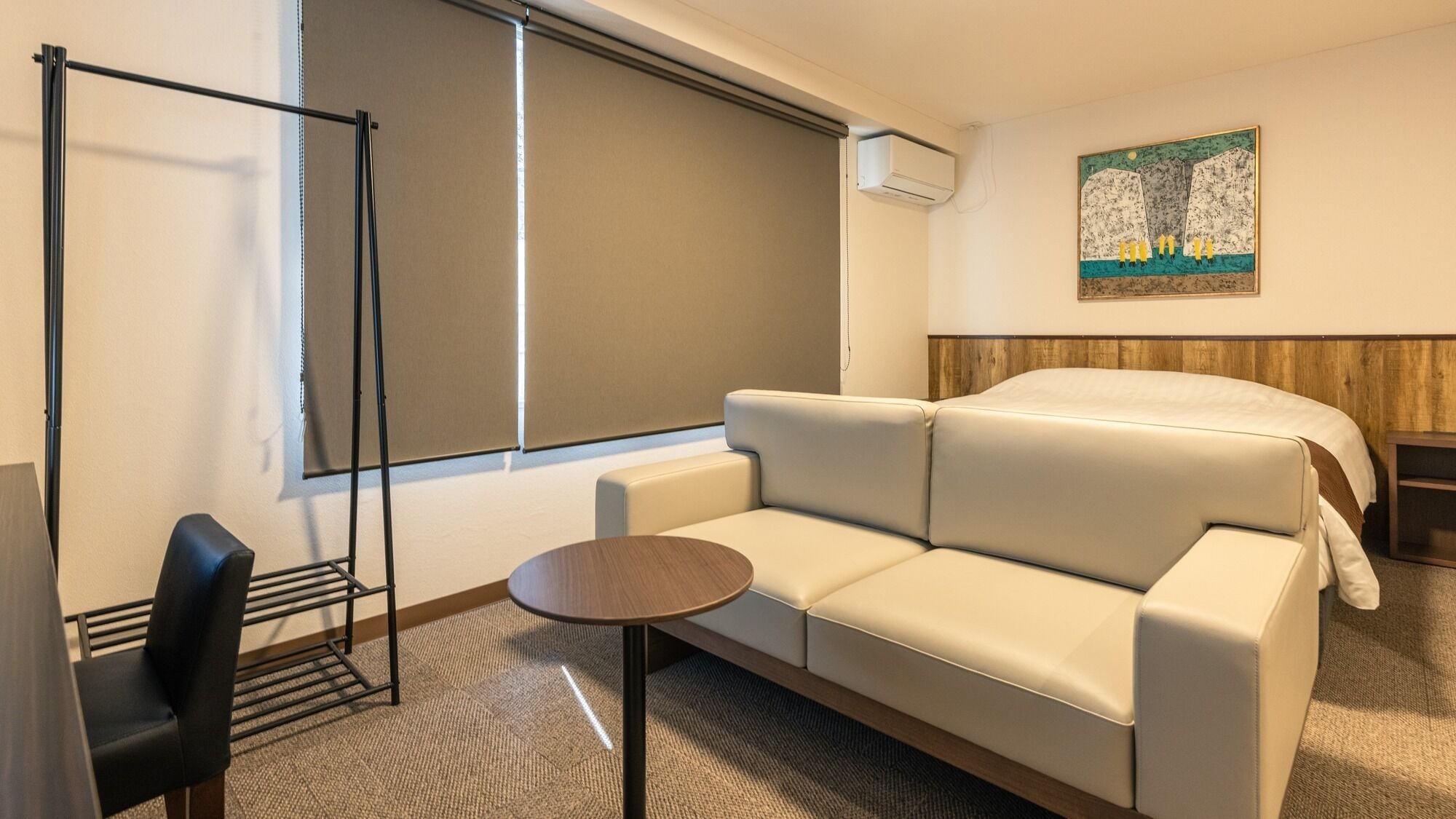 Quarto de hotel com sofá, mesa pequena, cama, janela com persianas, ar condicionado e um quadro na parede.