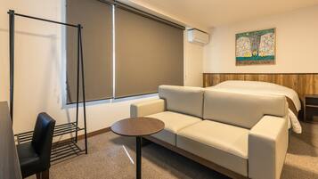 Quarto de hotel com sofá, mesa pequena, cama, janela com persianas, ar condicionado e um quadro na parede.