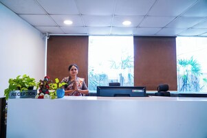 Uma receção com um letreiro, um computador, uma cadeira e plantas em vasos.