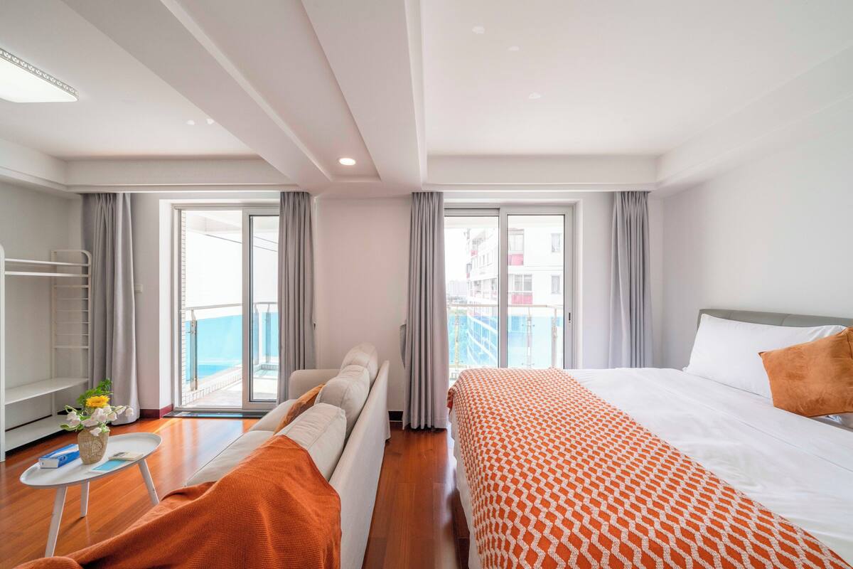 Quarto de hotel com cama, sofá, uma mesinha com um vaso e uma varanda com cortinas.