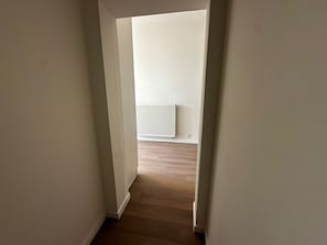 Um corredor estreito com soalho de madeira que leva a um quarto com radiador branco.